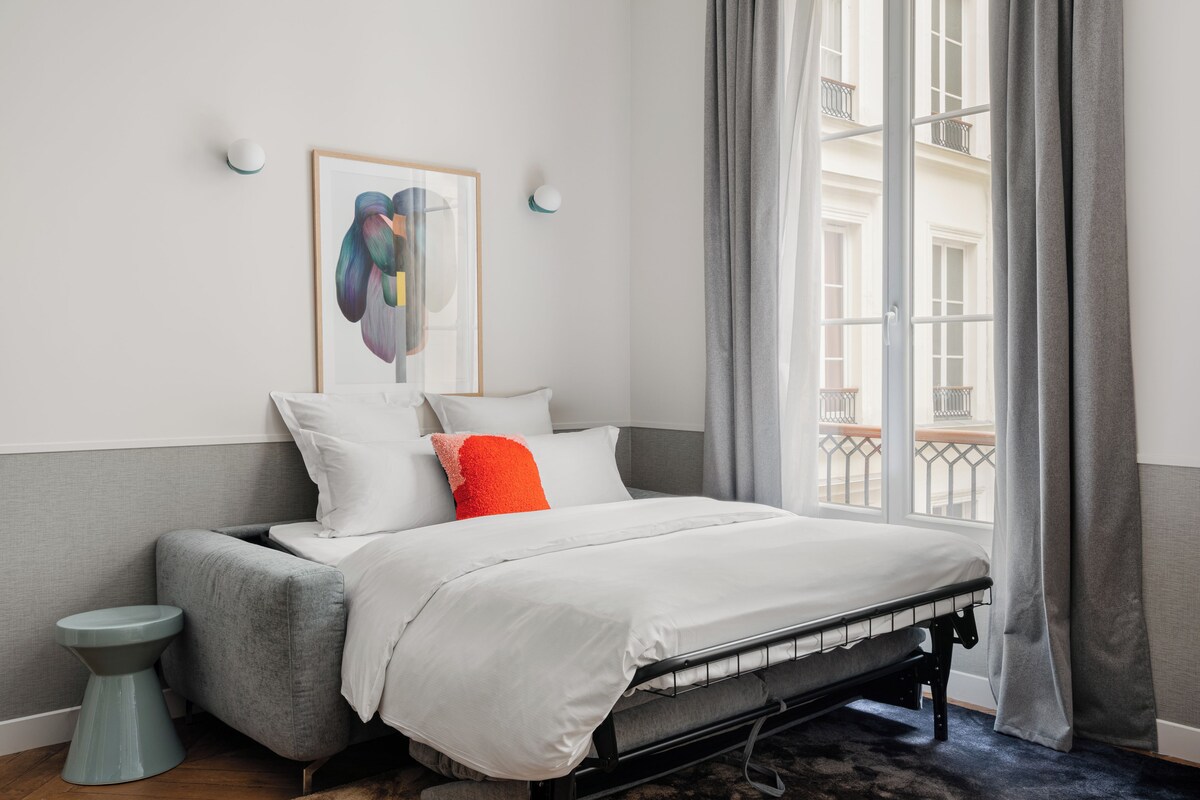 Quarto com cama, almofadas, mesa de cabeceira, obra de arte emoldurada e janela com cortinas.