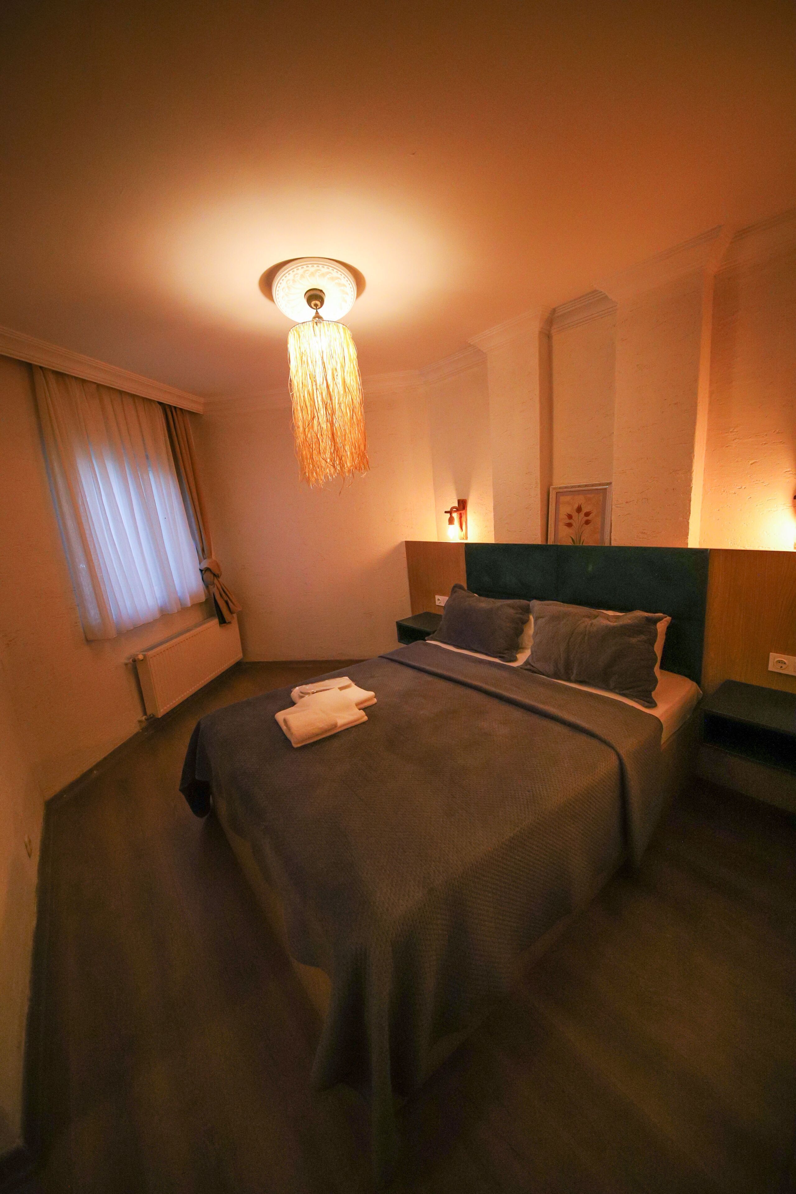Quarto de hotel com uma cama grande, luminária pendente e janela com cortinas.