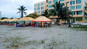Um hotel à beira-mar com diversas áreas de refeições sob toldos, palmeiras e uma praia arenosa.