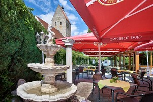 Um café ao ar livre com guarda-sóis vermelhos, uma fonte de pedra e uma igreja ao fundo.