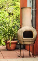 Um forno cerâmico grande sobre suporte de metal, uma planta em vaso e uma parede de tijolos.
