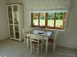 Uma área de refeições com uma mesa de madeira e cadeiras brancas, um armário de vidro com louças e uma janela com vista para o verde.