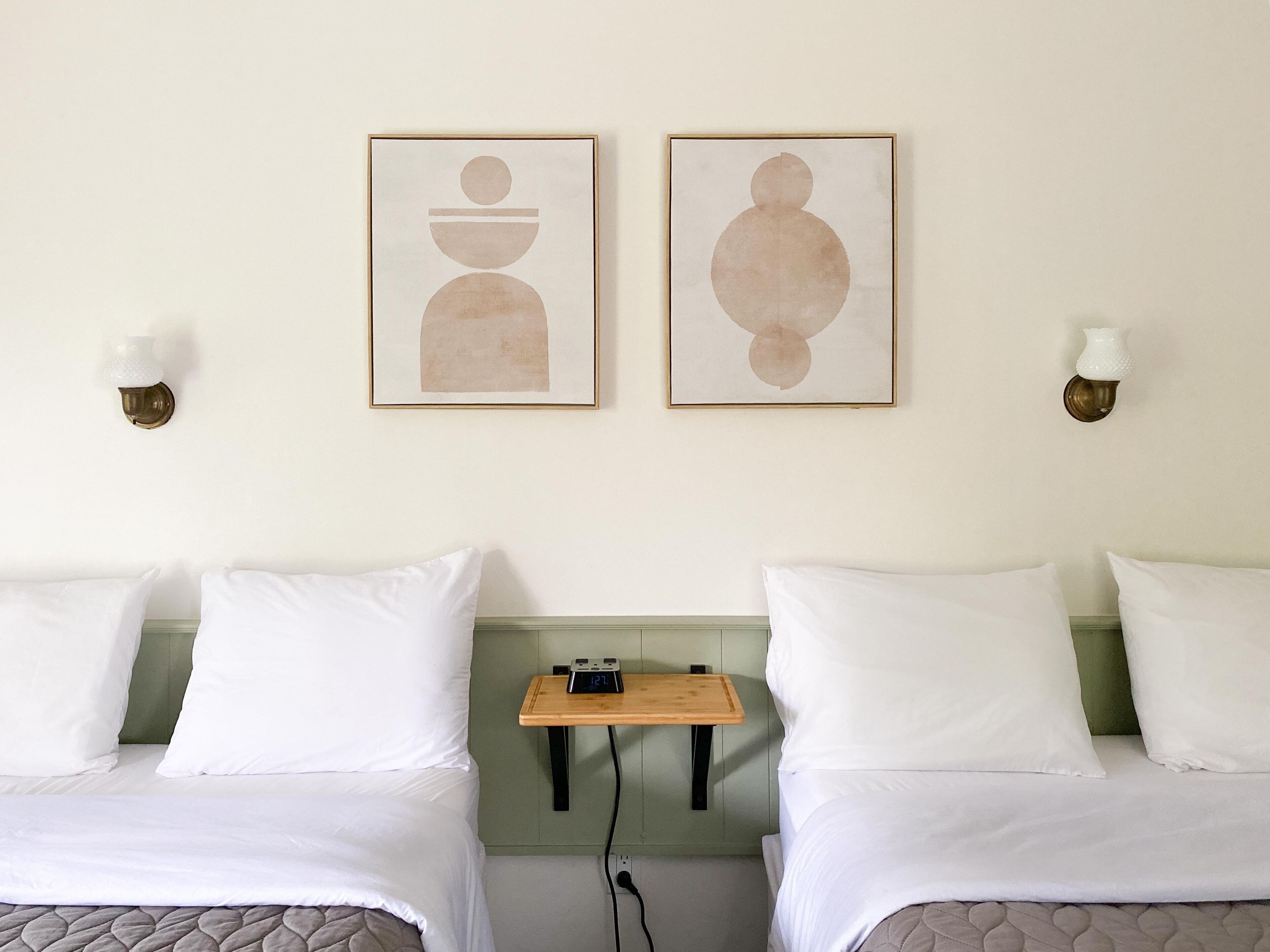 Quarto de hotel com duas camas, uma mesinha de cabeceira com um relógio e duas pinturas abstratas na parede.