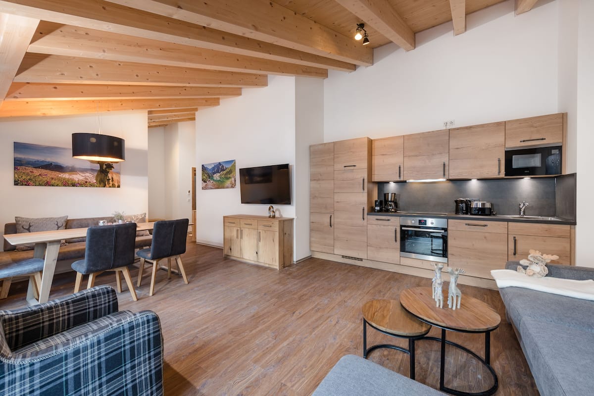 Uma cozinha moderna com armários de madeira, forno embutido e micro-ondas. Possui uma área de jantar com mesa e cadeiras, e uma sala de estar com sofá e mesinha de centro.