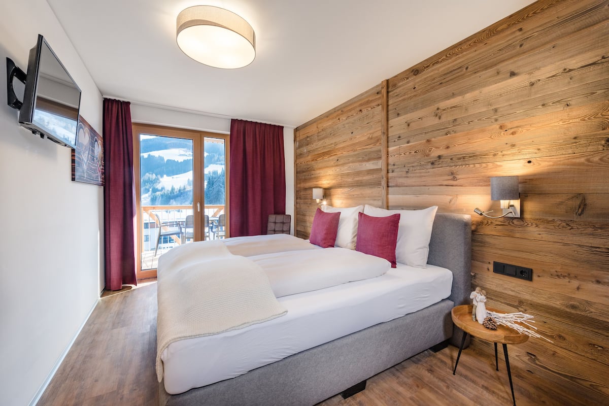 Quarto moderno com uma cama grande, paredes de painel de madeira, uma TV e vista para uma paisagem nevada.