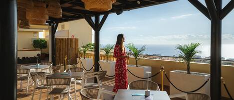 Um terraço com mobiliário de vime, uma mulher num vestido vermelho e vista para o mar.