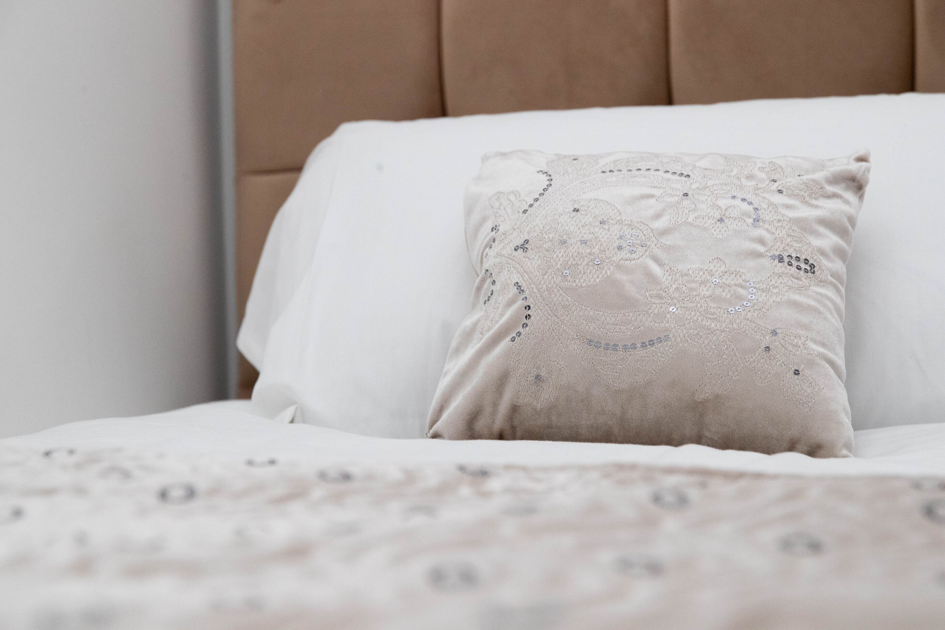 Uma cama com travesseiro bege com padrão floral e aplicações de lantejoulas, cobreleira branca com padrão discreto e cabeceira estofada marrom.