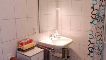 Casa de banho com uma pia branca, espelho e paredes revestidas de azulejos. Inclui uma sanita, um pequeno armário com uma toalha e um cortina de chuveiro com padrão floral.