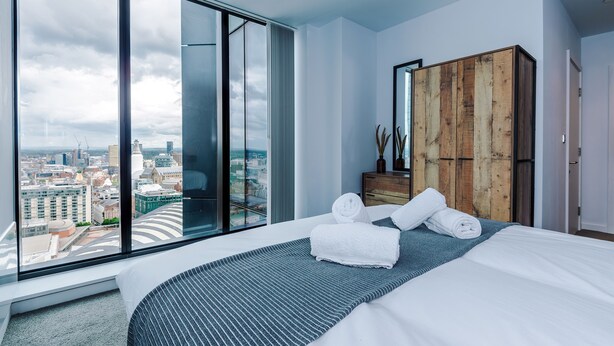 Quarto de hotel moderno com uma grande janela oferecendo vista da cidade, uma cama bem arrumada e um guarda-roupa de madeira.