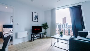 Quarto de hotel moderno com uma cama, uma TV exibindo 'NETFLIX', um vaso de planta, uma mesa de jantar e uma janela grande com vista para a cidade.