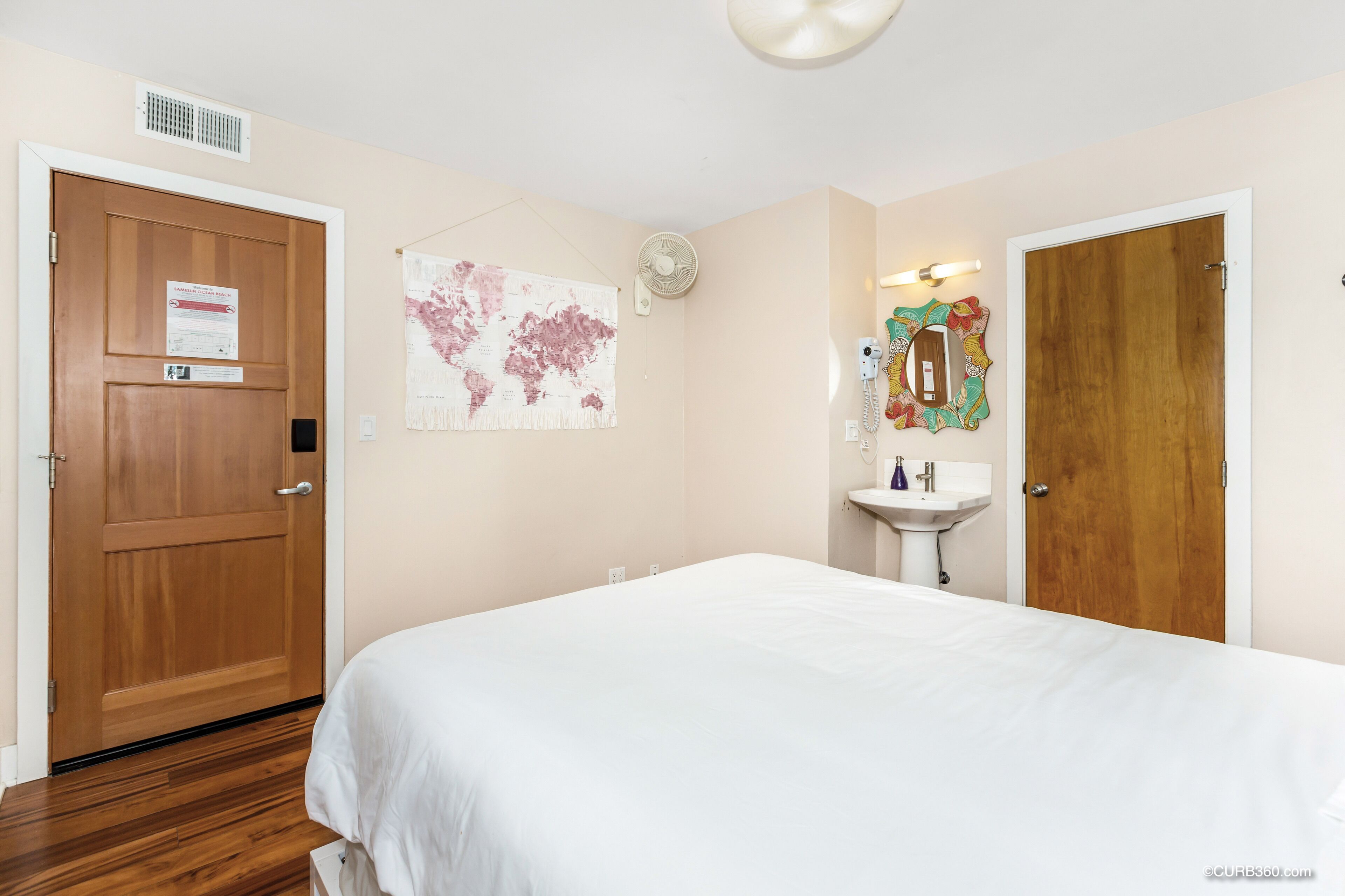Quarto de hotel com porta de madeira, cama com roupa de cama branca, mapa fixado na parede, um ventilador, pia com espelho e luminária.