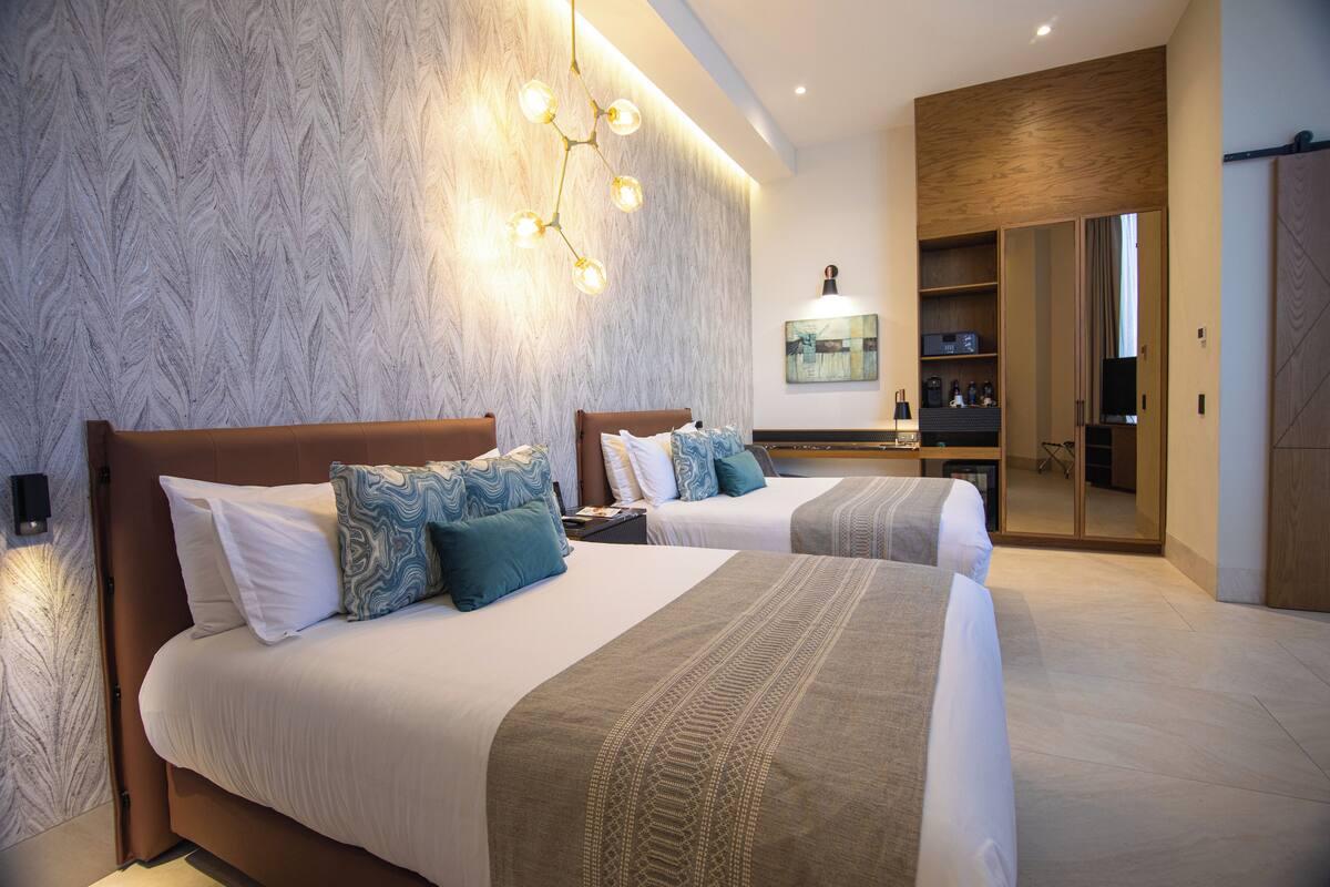Quarto de hotel com duas camas, guarda-roupa de madeira, espelho e luminária na parede.