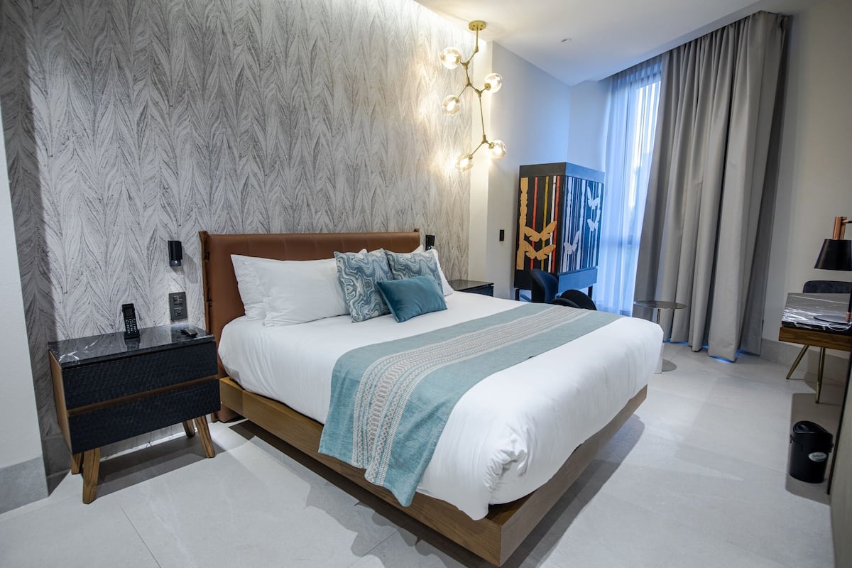 Quarto de hotel moderno com uma cama grande, mesa de cabeceira, escrivaninha e luminária na parede.