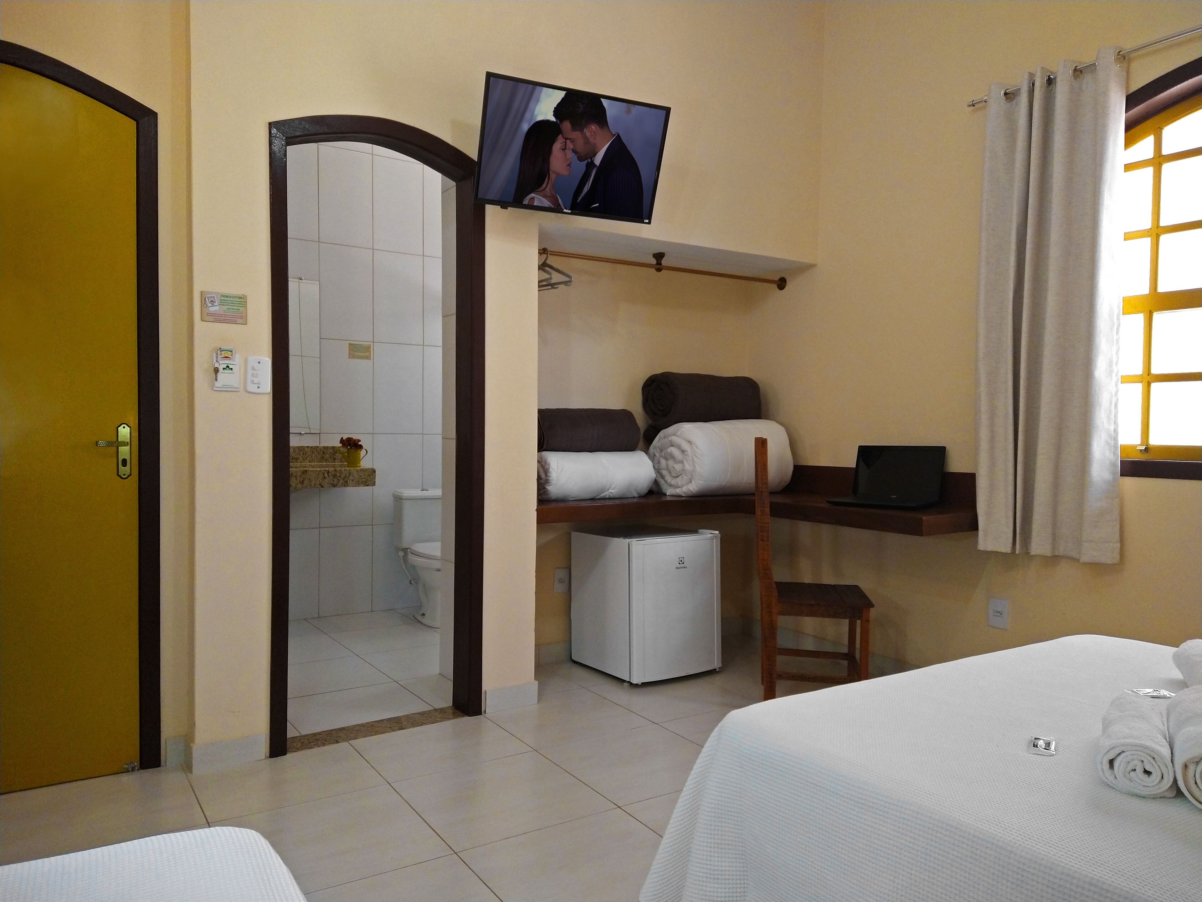Quarto de hotel com cama, televisão fixada na parede, mesa pequena, cadeira, prateleira com toalhas e janela com cortinas.