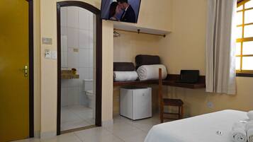 Quarto de hotel com cama, televisão fixada na parede, mesa pequena, cadeira, prateleira com toalhas e janela com cortinas.
