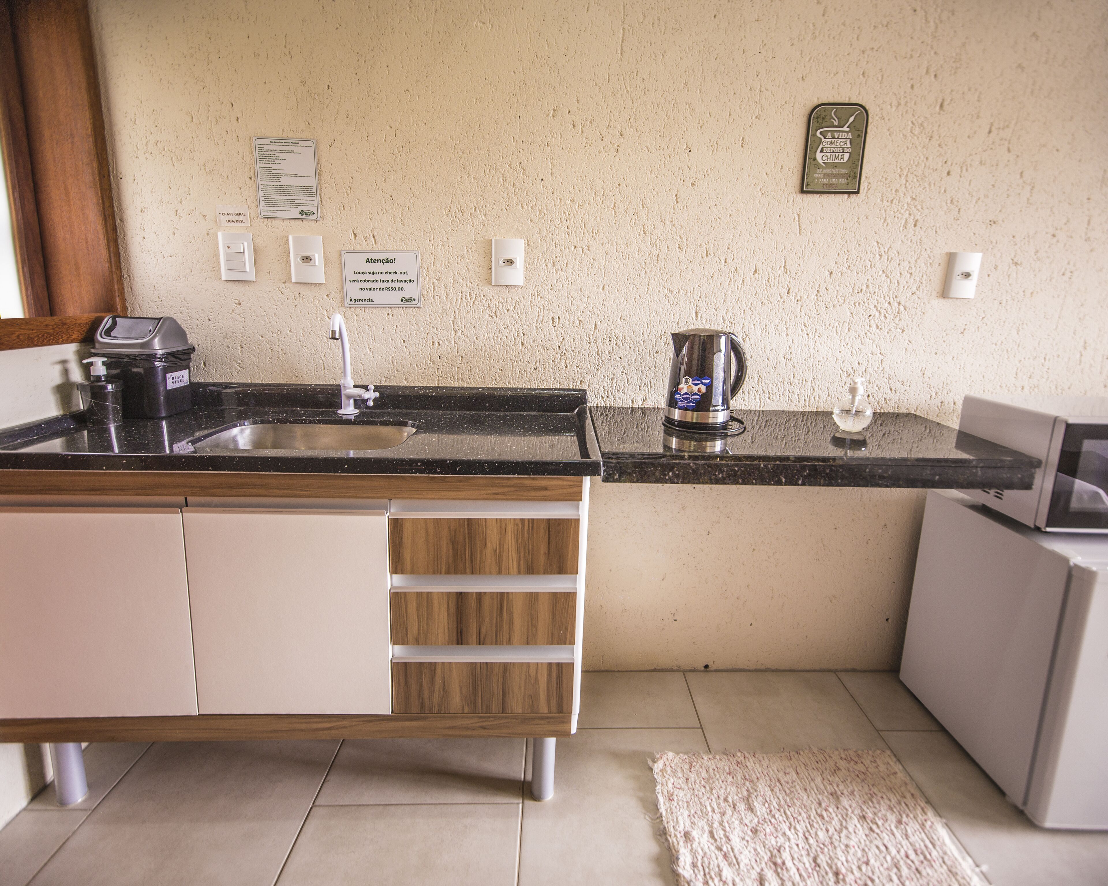 Cozinha compacta com pia, cafeteira e micro-ondas. Há armários com puxadores de madeira e um tapete pequeno no chão.