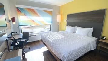 Quarto de hotel com uma cama grande, uma escrivaninha com cadeira, uma janela com vista e um quadro colorido na parede.