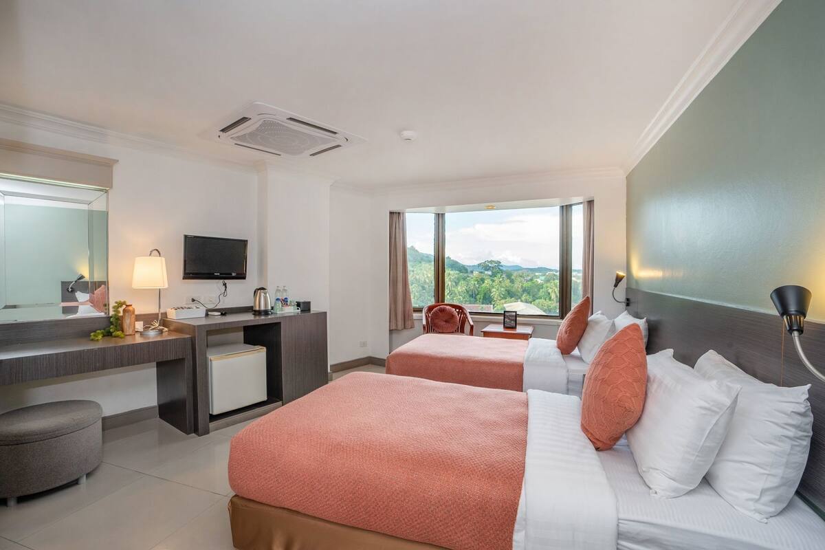 Quarto de hotel com duas camas, uma escrivaninha, televisão e janela com vista.