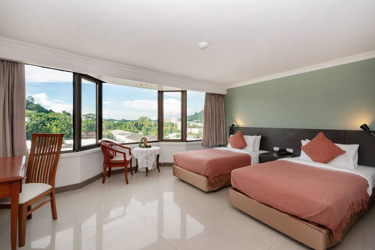 Quarto de hotel com duas camas, uma mesa de jantar, cadeiras, uma mesinha com toalha branca e grandes janelas com vista para áreas verdes e edifícios.