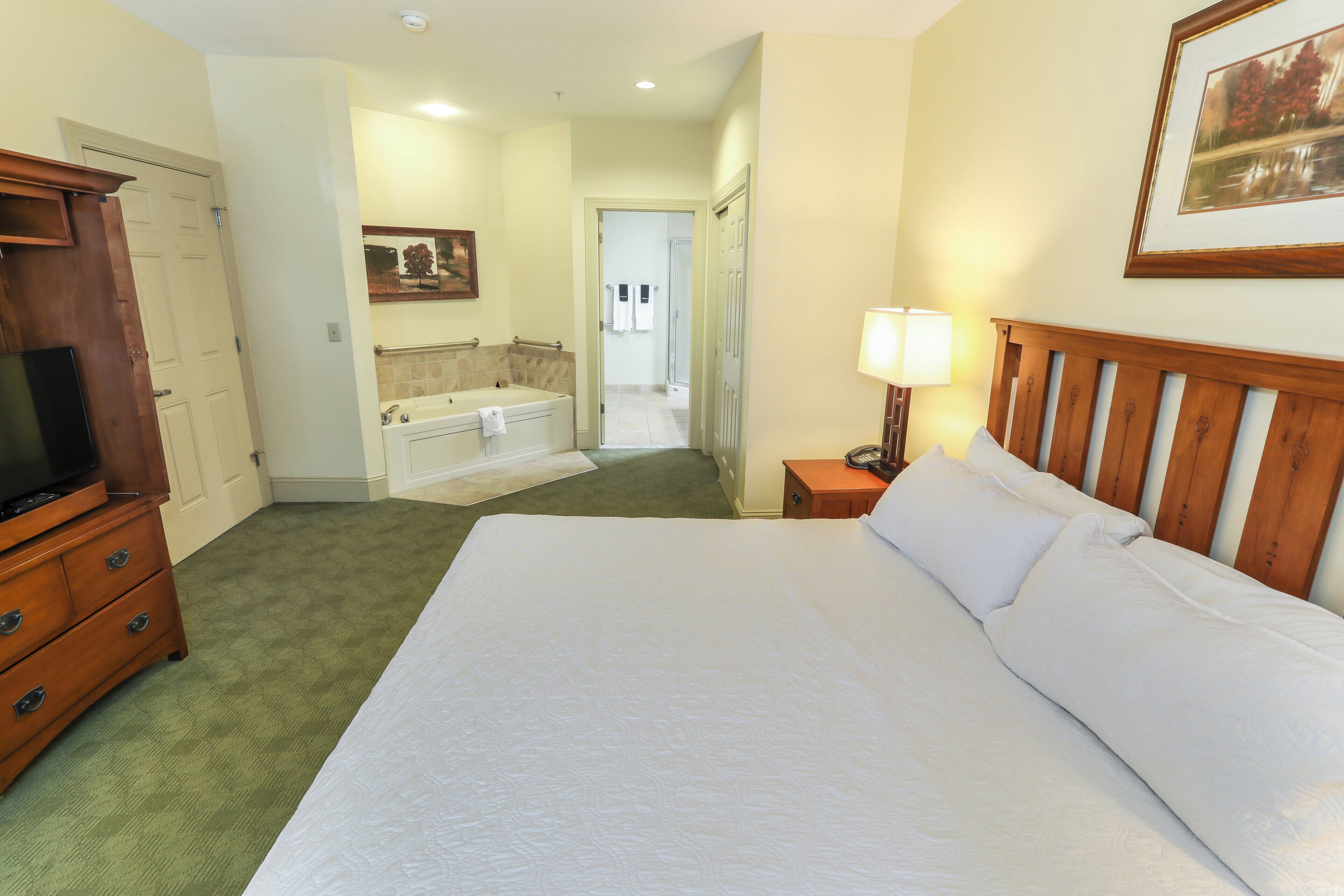 Quarto de hotel com uma cama grande, cómoda de madeira, televisão, banheiro com banheira e mesa de cabeceira com abajur.