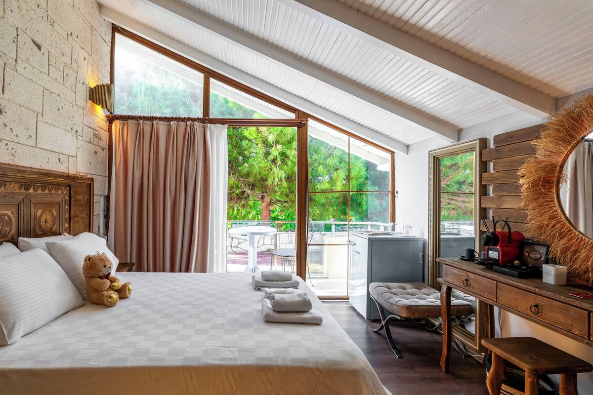 Quarto com cama grande, escrivaninha de madeira, cadeira, cômoda e grande janela com vista para árvores.