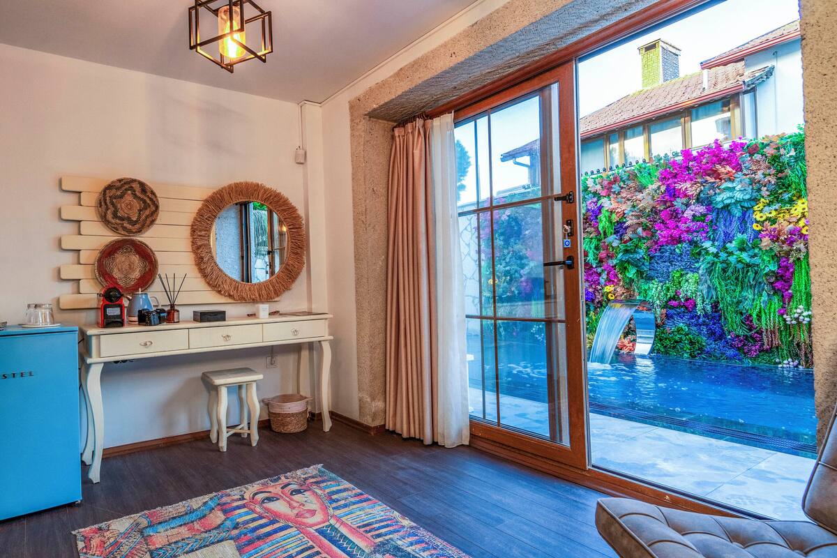 Um quarto com uma escrivaninha de madeira, uma cómoda azul, um espelho e vista para um jardim colorido através de uma porta de vidro.