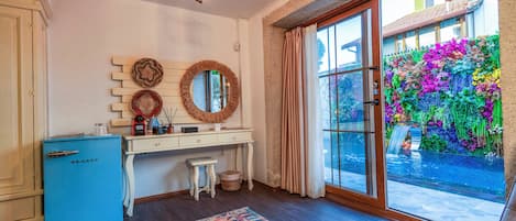Um quarto com uma escrivaninha de madeira, uma cómoda azul, um espelho e vista para um jardim colorido através de uma porta de vidro.