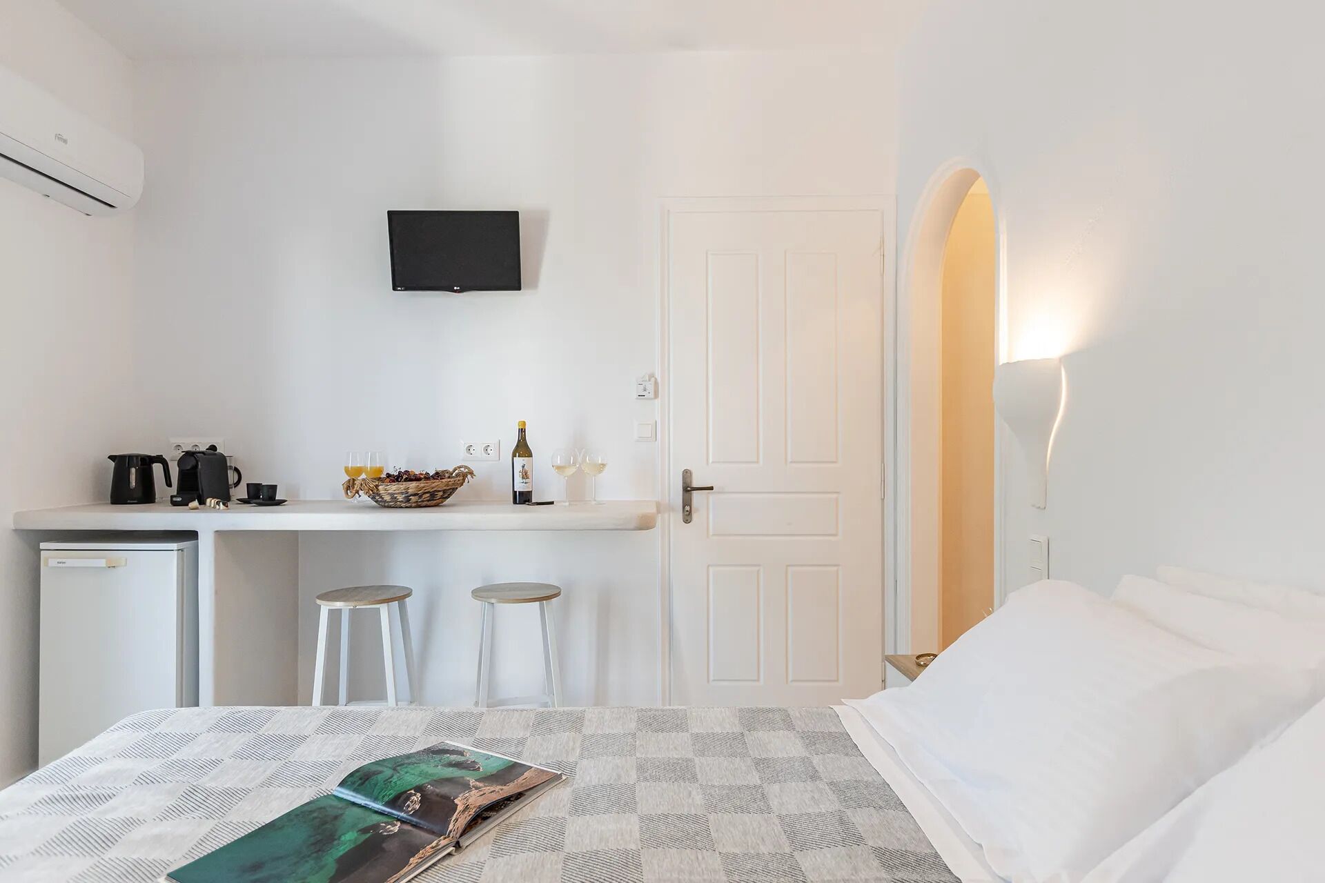 Quarto de hotel com cama, pequeno espaço de cozinha, televisão presa à parede e porta com luz.