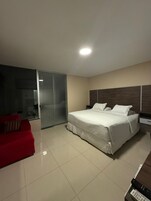Quarto de hotel com uma cama, um sofá vermelho, um guarda-roupa com porta de vidro e uma cabeceira de madeira.