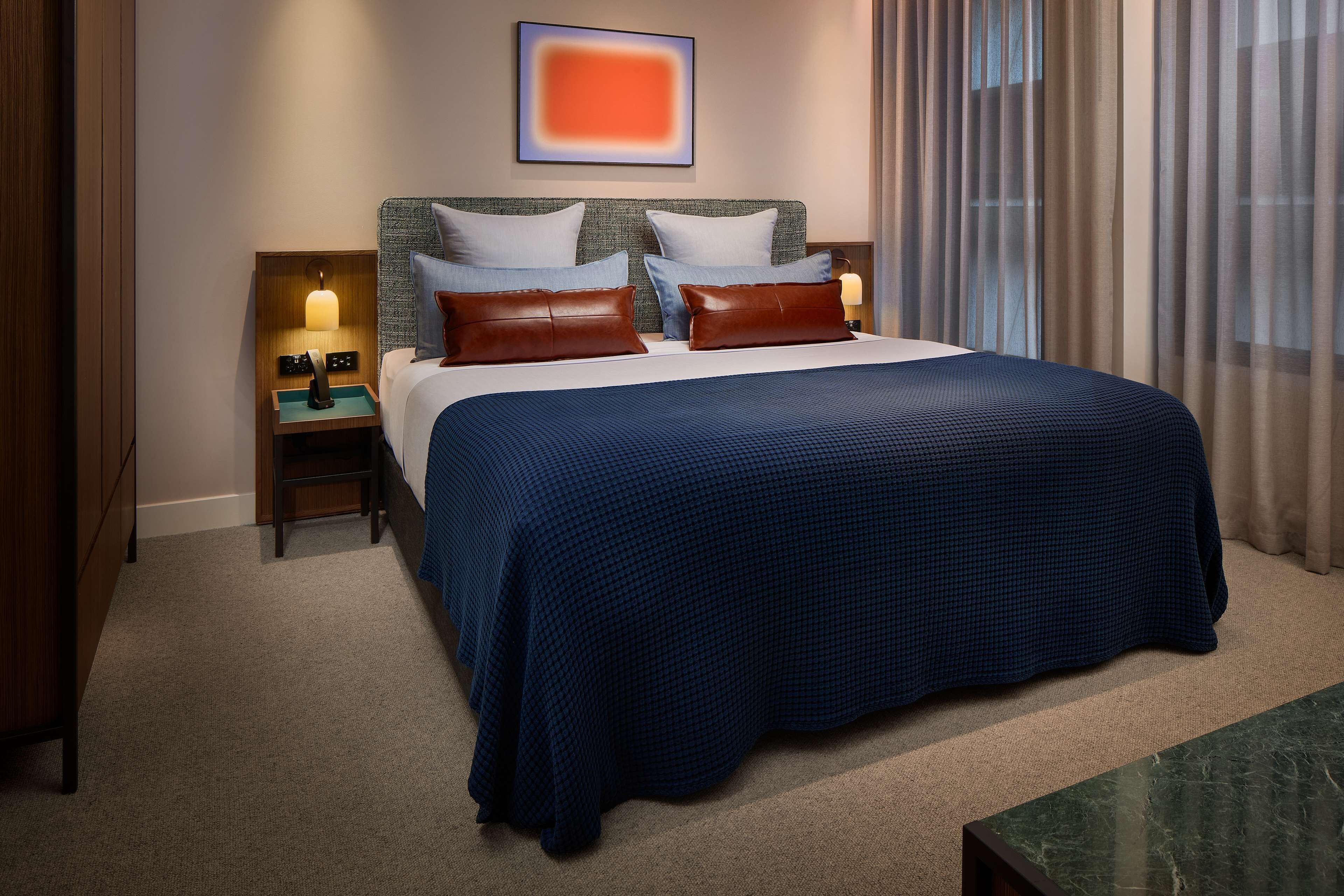 Quarto de hotel com uma cama grande, duas mesas de cabeceira, uma luminária de parede e uma janela com cortinas.