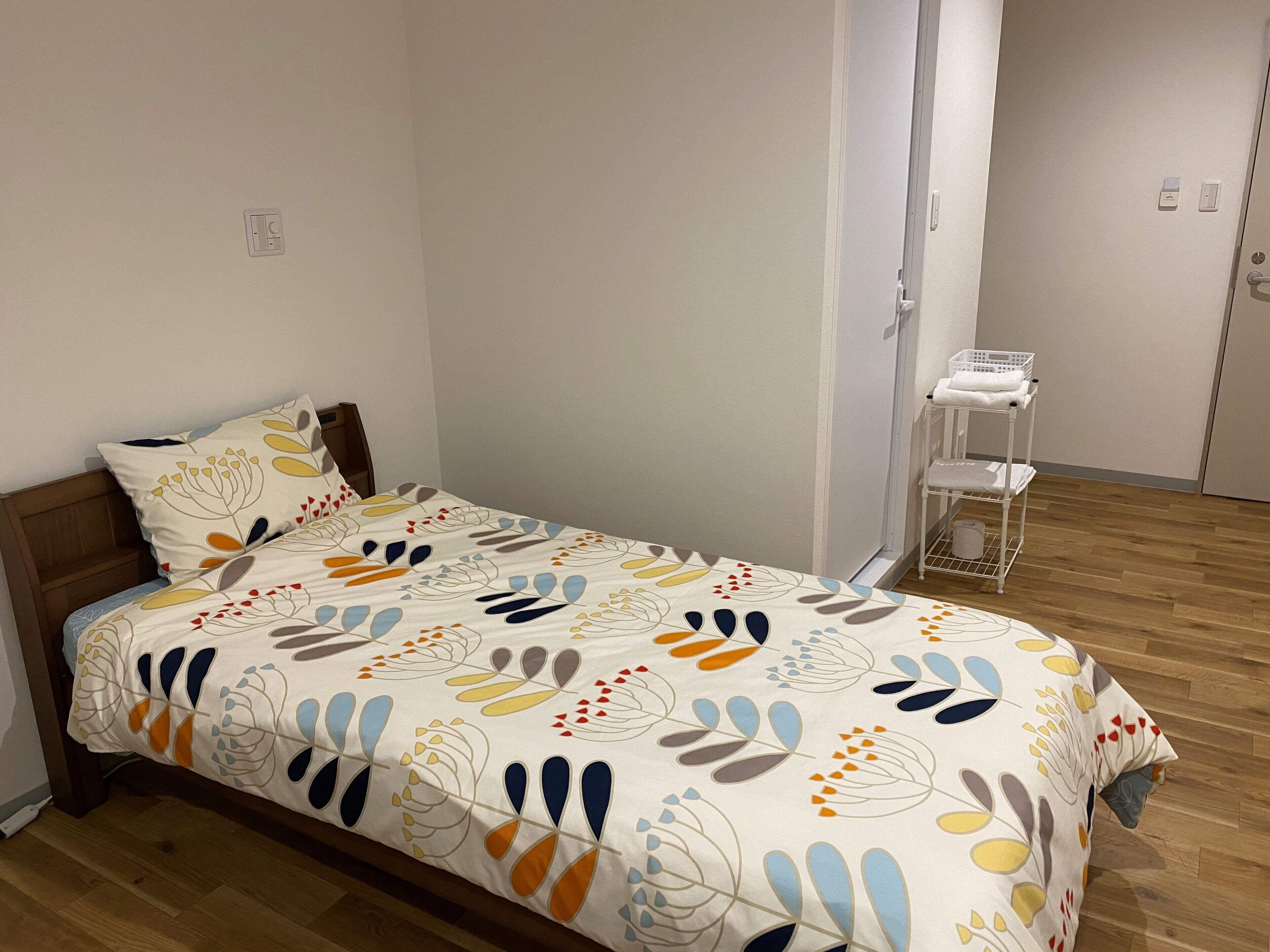 Uma cama de solteiro com colcha estampada, cabeceira de madeira, parede branca, uma porta e um cesto de roupa branco.
