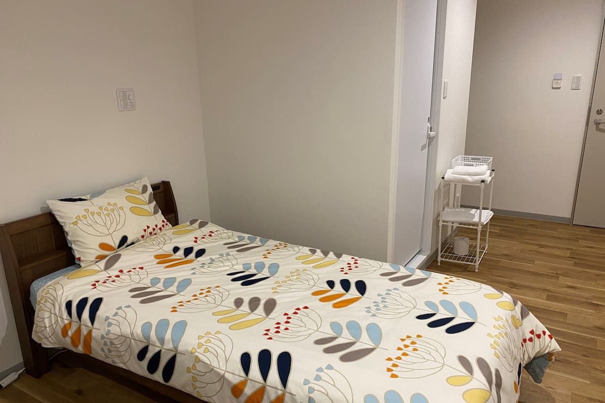 Uma cama de solteiro com colcha estampada, cabeceira de madeira, parede branca, uma porta e um cesto de roupa branco.