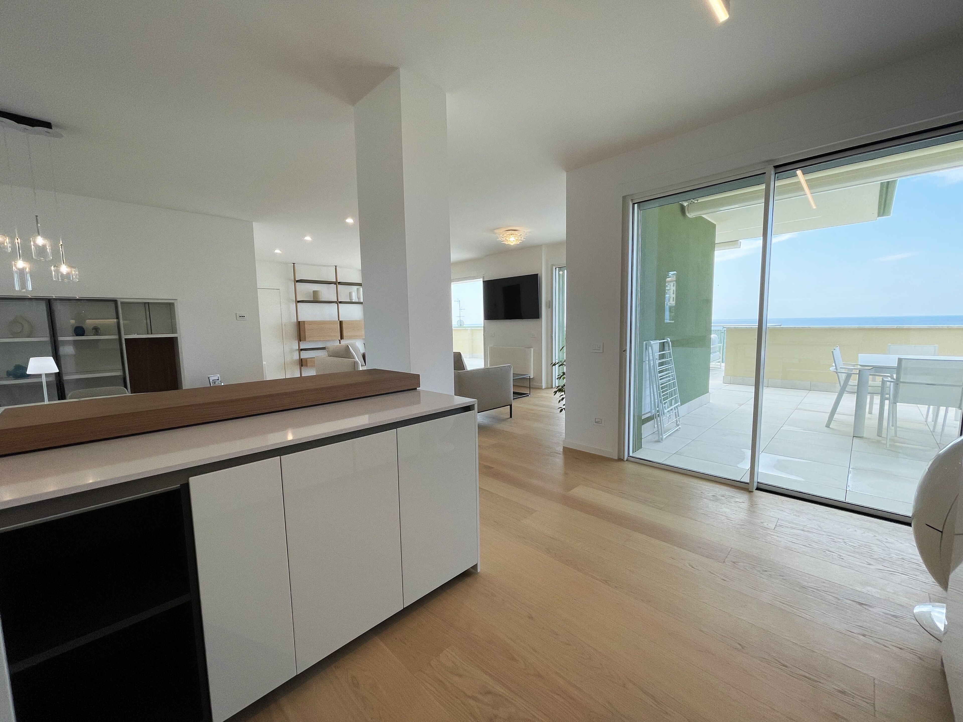 Cozinha moderna com bancada de madeira e armários brancos, área de jantar com mesa e cadeiras, e uma grande porta de vidro deslizante que dá para uma varanda com vista para o mar.