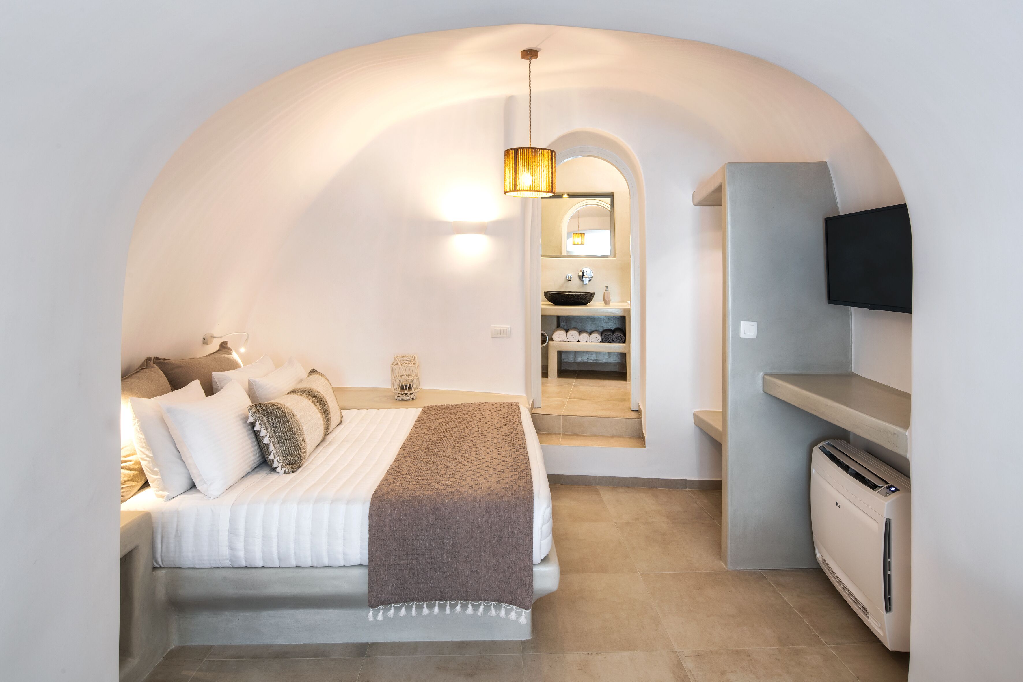 Quarto com cama, mesa de cabeceira, abajur e televisão fixada na parede.