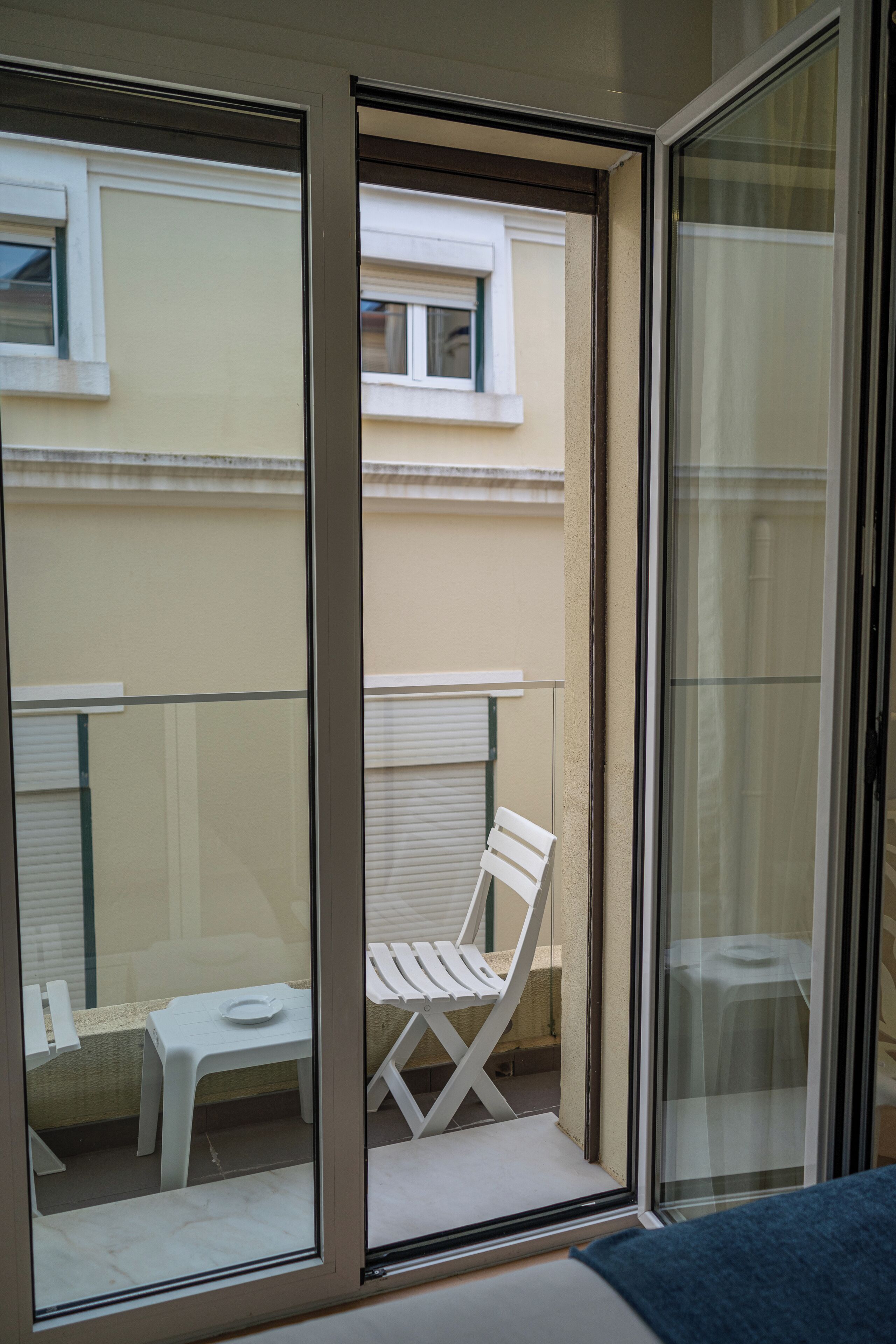 Uma varanda com cadeira e mesa brancas, porta de vidro e vista para um prédio com janelas.