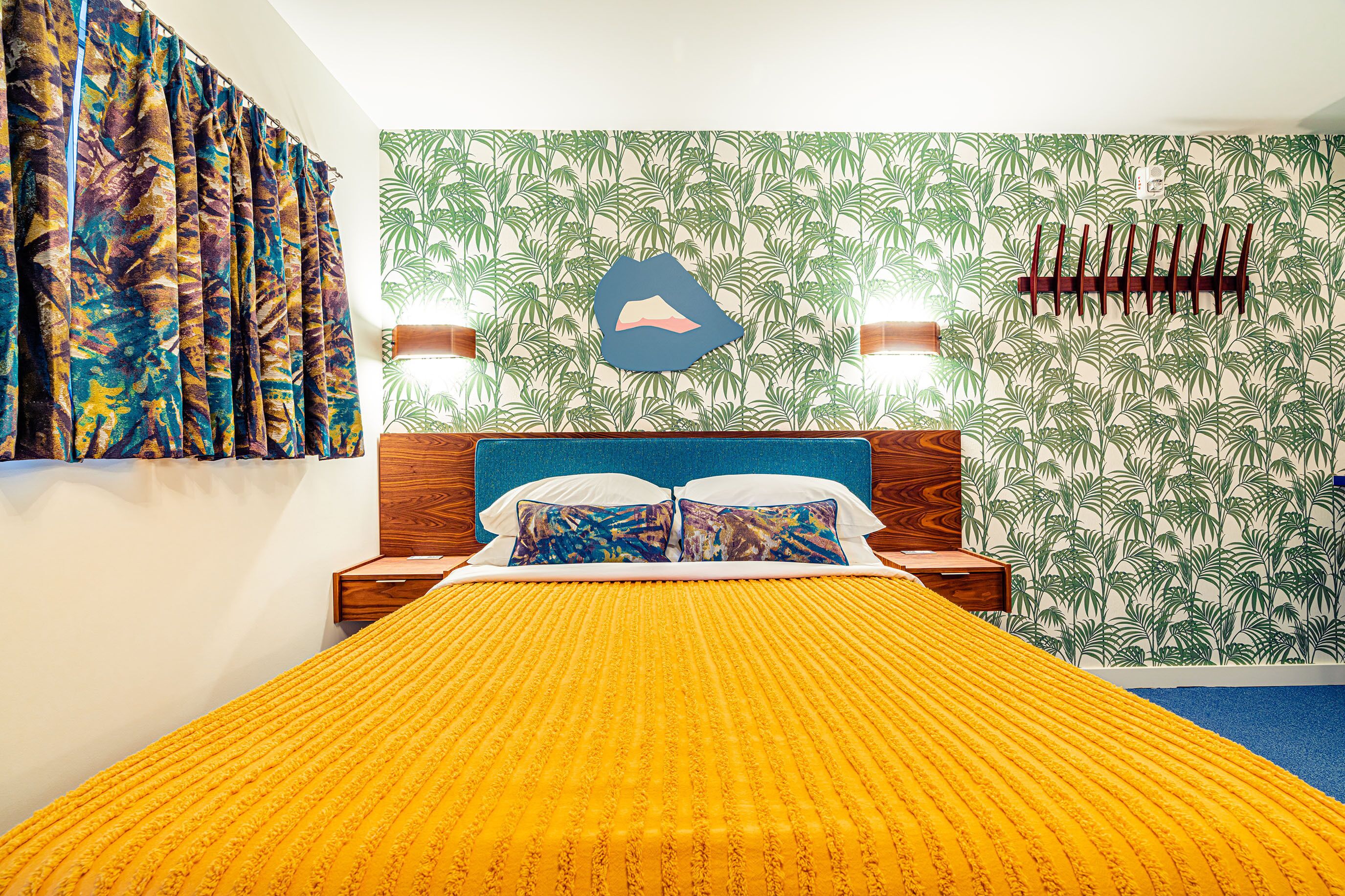 Quarto com cama, mesa de cabeceira, televisão na parede e papel de parede tropical.