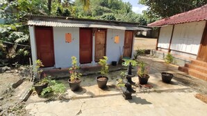 Um pequeno edifício térreo, com telhado de metal corrugado e portas de madeira. Diversas plantas em vasos estão dispostas em frente ao edifício, e uma pequena estrutura de pedra à esquerda.