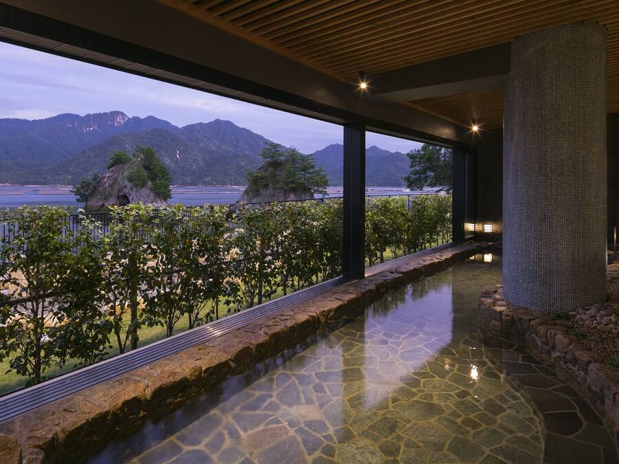 Interior moderno com piscina revestida em pedra, vista para montanhas e lago, e teto de madeira.