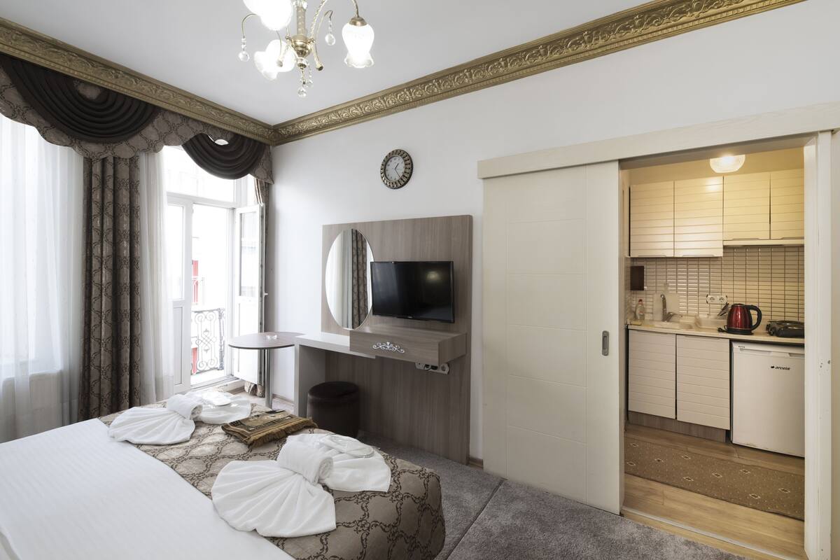 Quarto de hotel com cama, televisão, uma pequena mesa, espelho e vista para a cozinha.