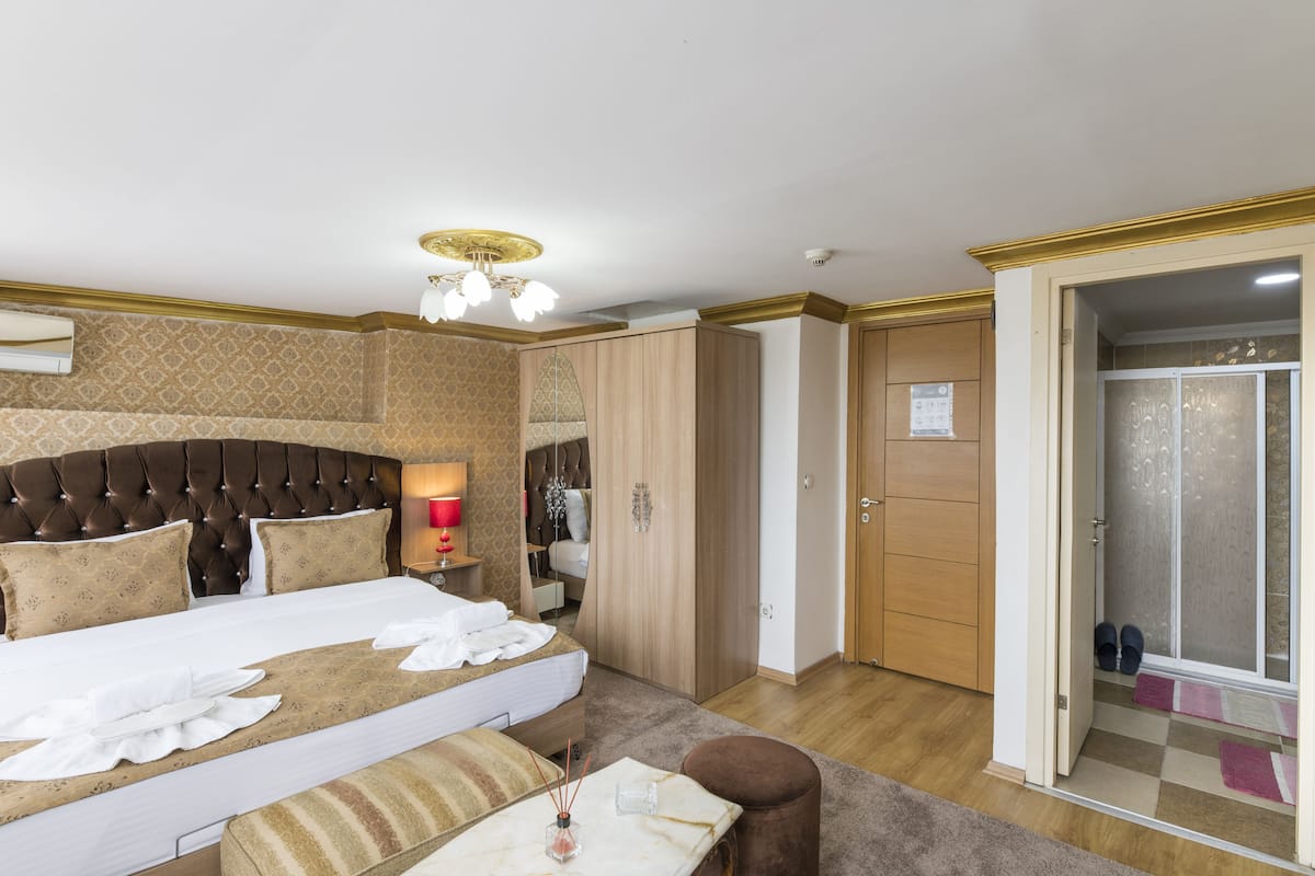 Quarto de hotel com uma cama grande, uma mesinha de cabeceira com luminária, um guarda-roupa, banheiro com chuveiro e uma porta que dá para outro quarto.