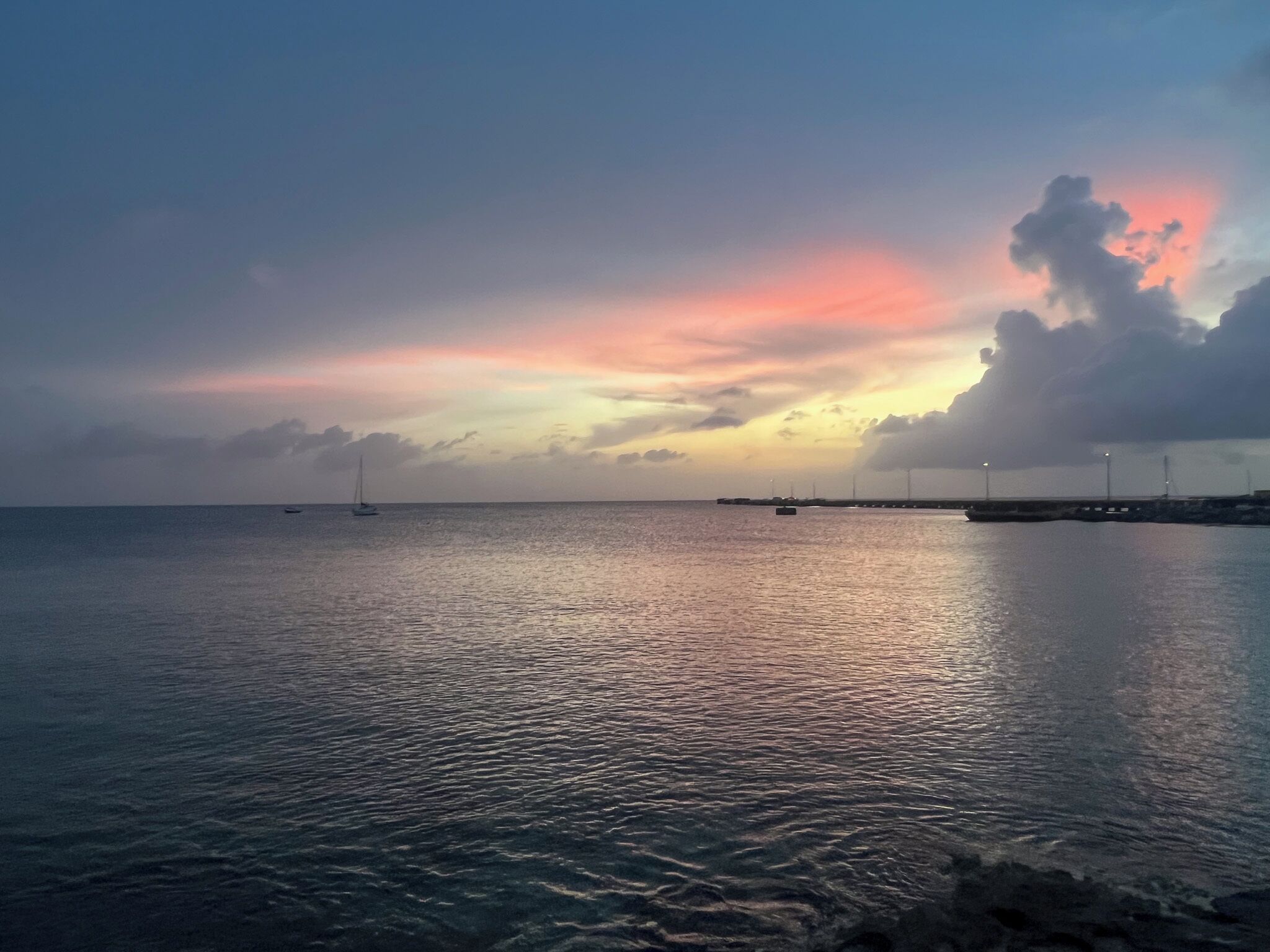 Mar calmo sob um céu de pôr do sol, um píer e um veleiro.
