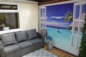Quarto com sofá de vime, tapete estampado, lanterna decorativa e um mural de uma praia tropical.