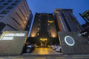 Entrada de hotel com letreiro de neon, carros estacionados e fachada de prédio moderno.