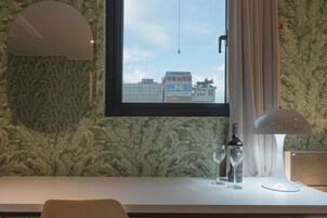 Quarto de hotel com uma janela grande, uma mesa com uma garrafa e copos, e uma cortina.