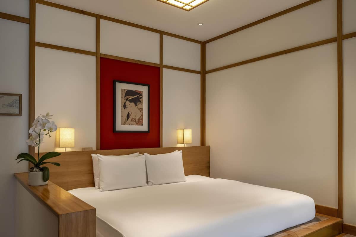 Quarto de hotel com cama, abajures, parede com detalhes em vermelho e uma obra de arte emoldurada.