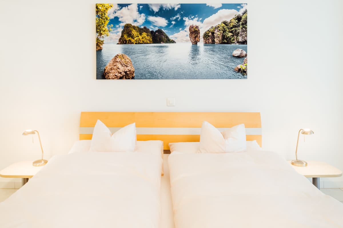 Quarto de hotel com duas camas, uma janela ampla com vista para um lago e montanhas, e luminárias de cabeceira.