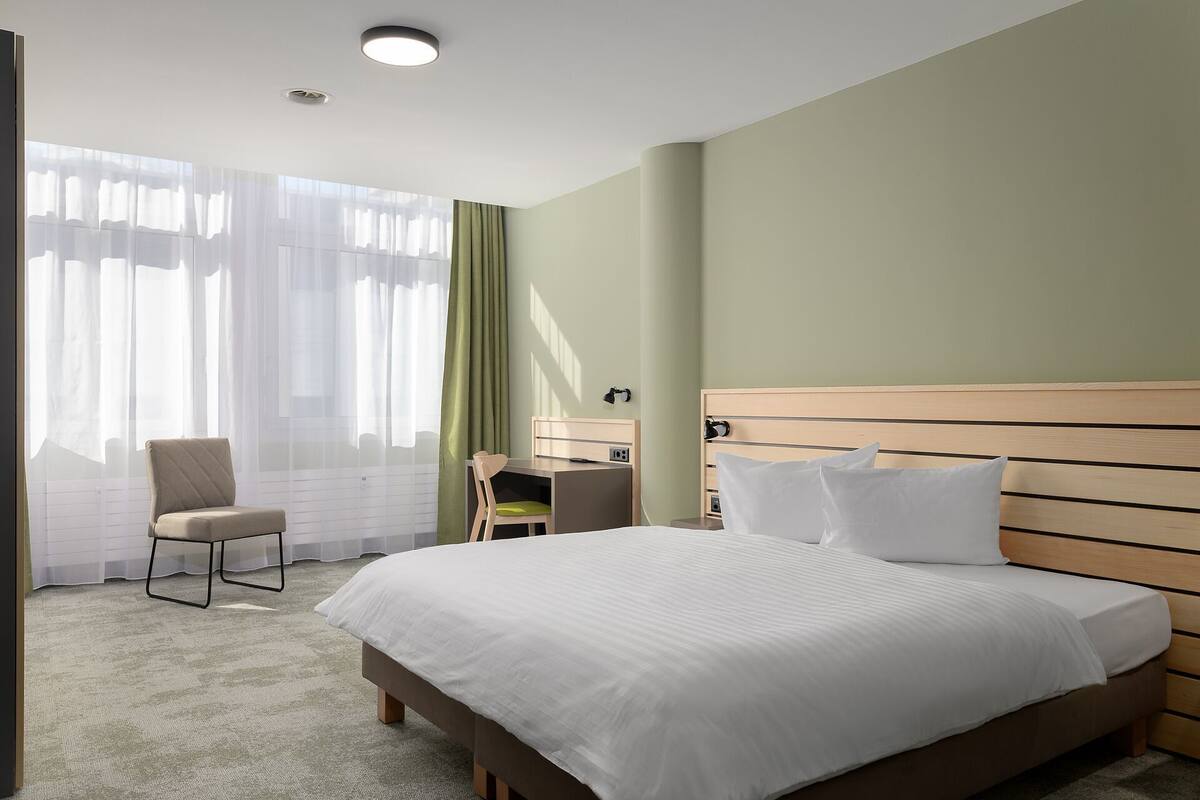 Quarto de hotel com uma cama grande, uma escrivaninha, uma cadeira e uma janela com cortinas finas.