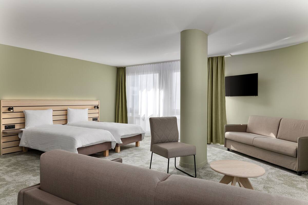 Quarto de hotel com duas camas, um sofá, uma poltrona, uma mesa pequena e uma TV fixada na parede.