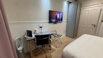 Quarto de hotel com cama, escrivaninha e cadeira, televisão fixada na parede e porta.