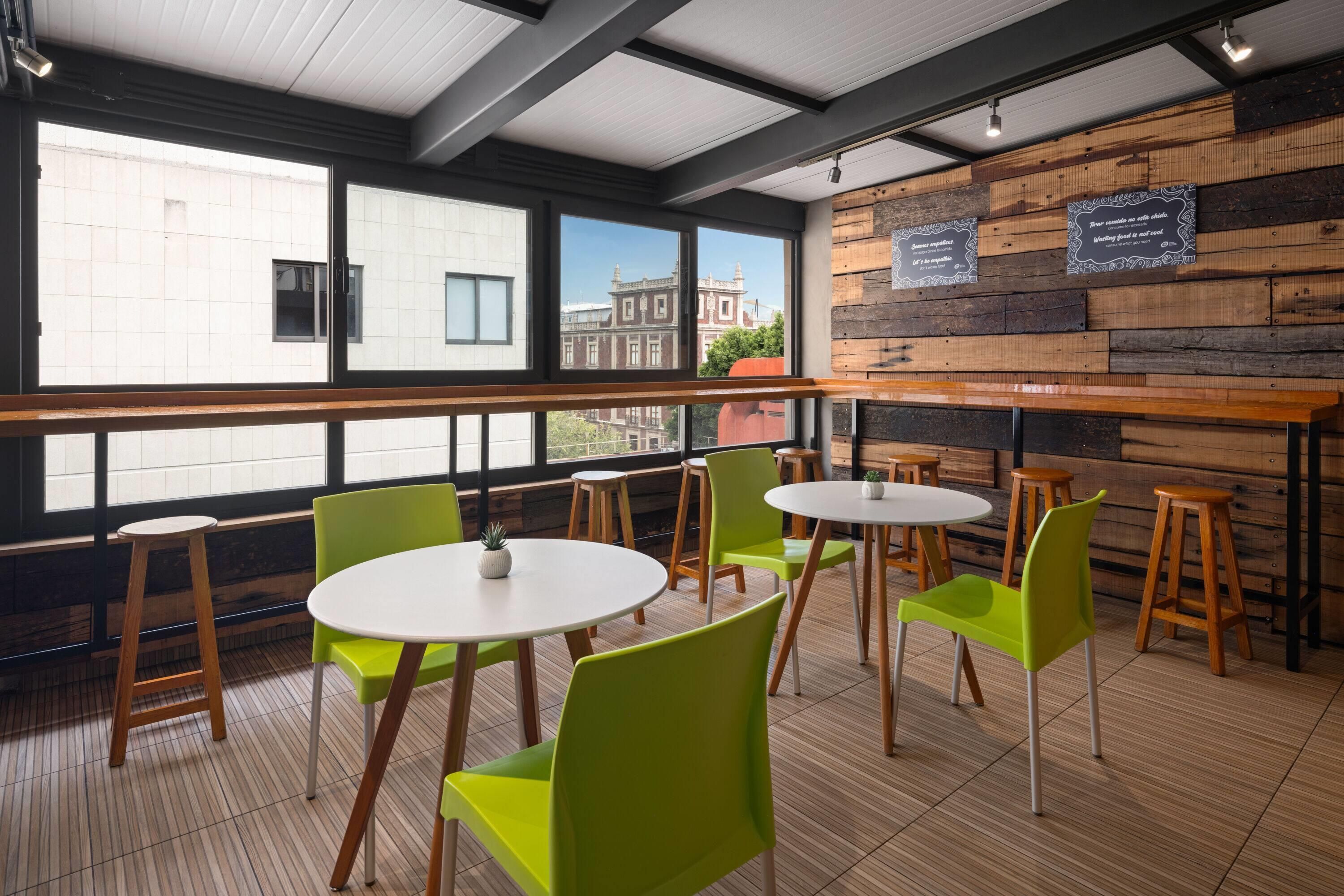 Interior de café moderno com mesas redondas e cadeiras verdes, grandes janelas com vista para edifícios e uma parede de madeira com mensagens em lousa.